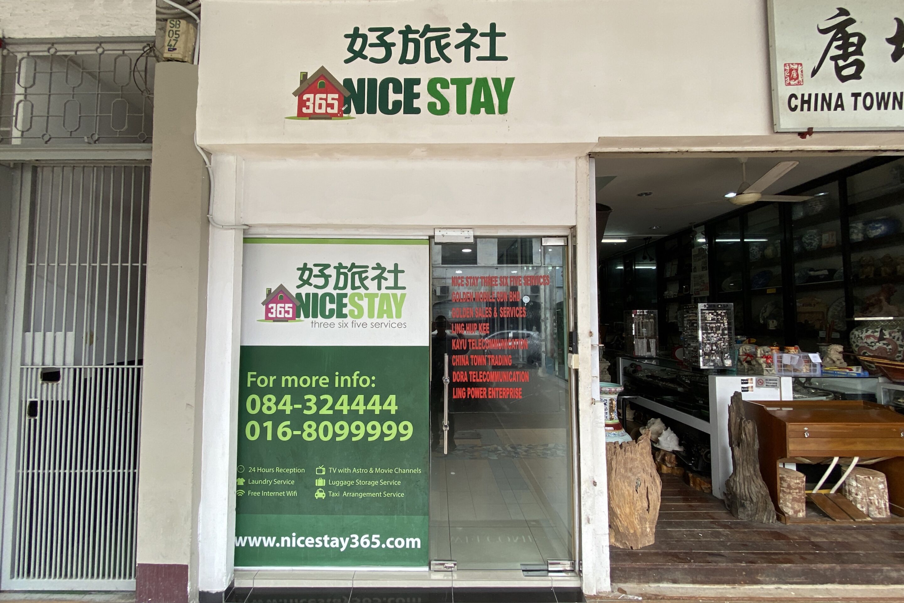 Entrada de um hotel com uma placa exibindo serviços como recepção 24 horas, lavanderia e TV com canais Astro e filmes.