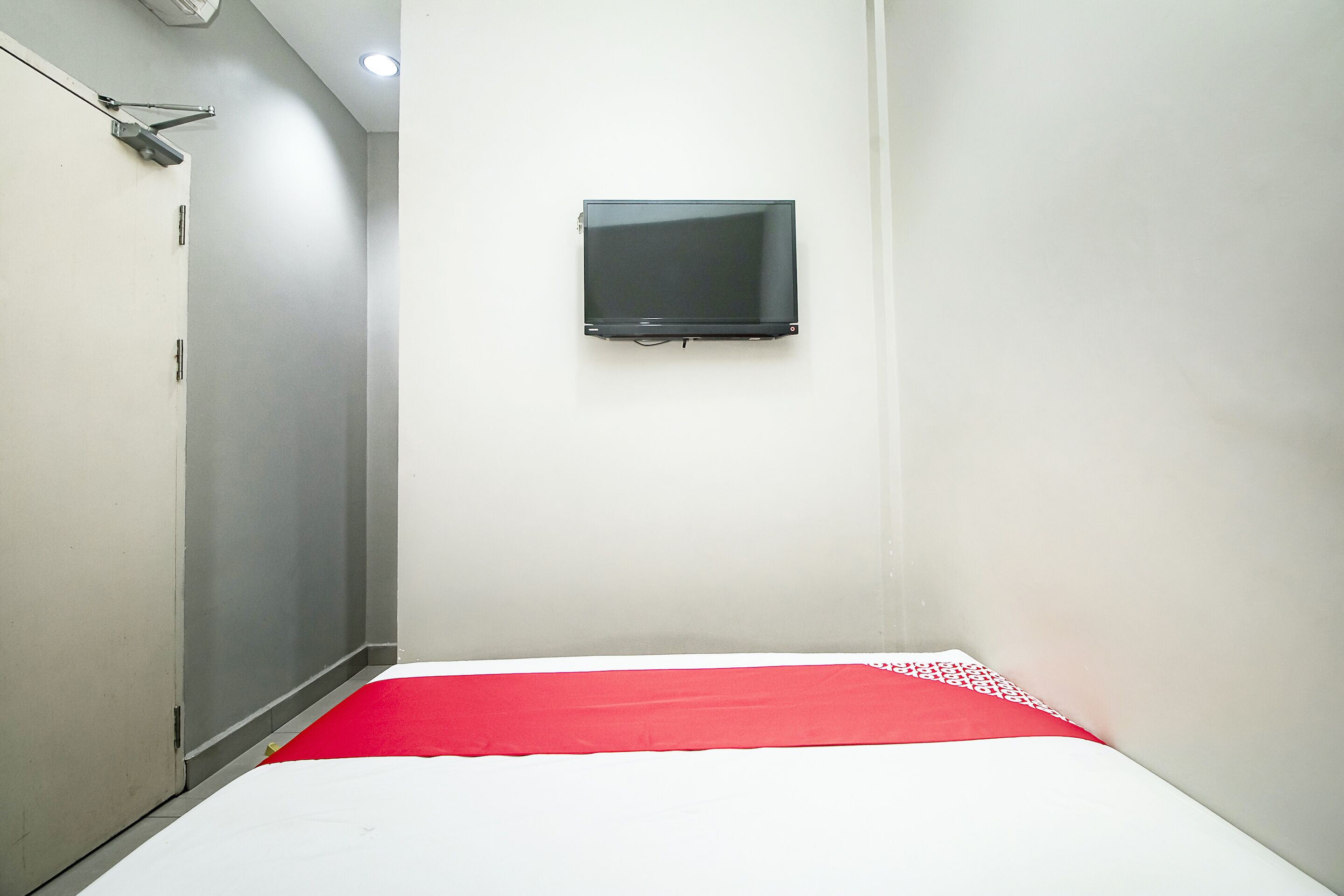 Um quarto pequeno e estreito, com uma cama, uma televisão na parede e uma porta.
