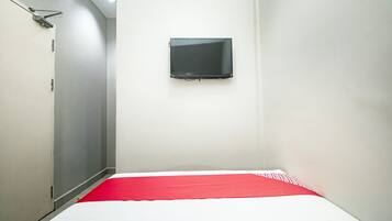 Um quarto pequeno e estreito, com uma cama, uma televisão na parede e uma porta.