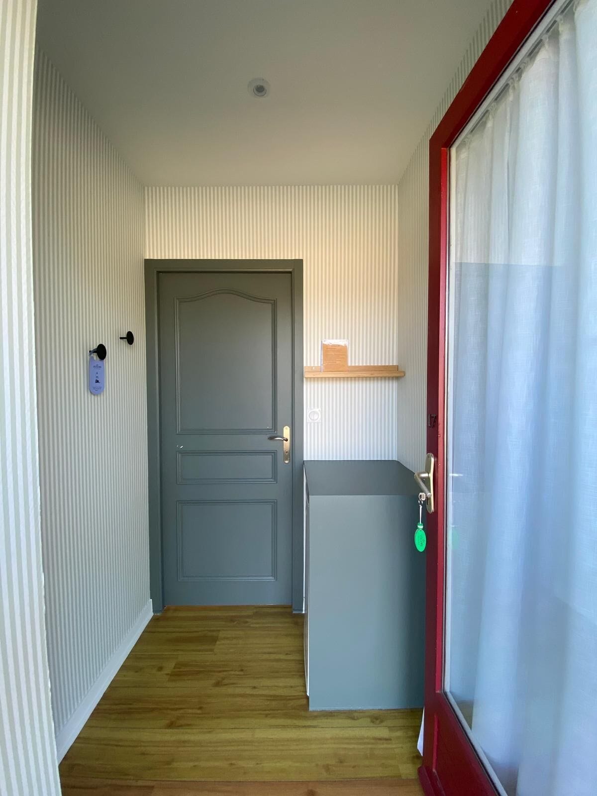 Um corredor estreito com piso de madeira, uma porta cinza e uma pequena cozinha compacta com geladeira e pia.