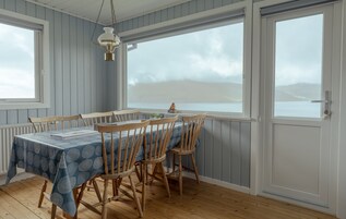 Uma sala de jantar com uma mesa posta para quatro pessoas, cadeiras de madeira, toalha de mesa estampada e vista para o mar através de grandes janelas.