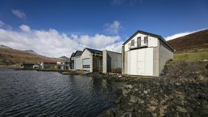 Uma fileira de edifícios à beira da água, com terreno rochoso e montanhas ao fundo.