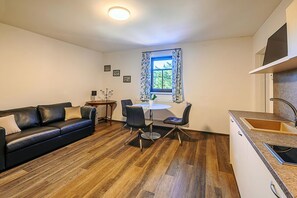 Um espaço de estar compacto com sofá de couro preto, mesa de jantar com quatro cadeiras, uma pequena mesa lateral e uma cozinha compacta com pia e bancada.