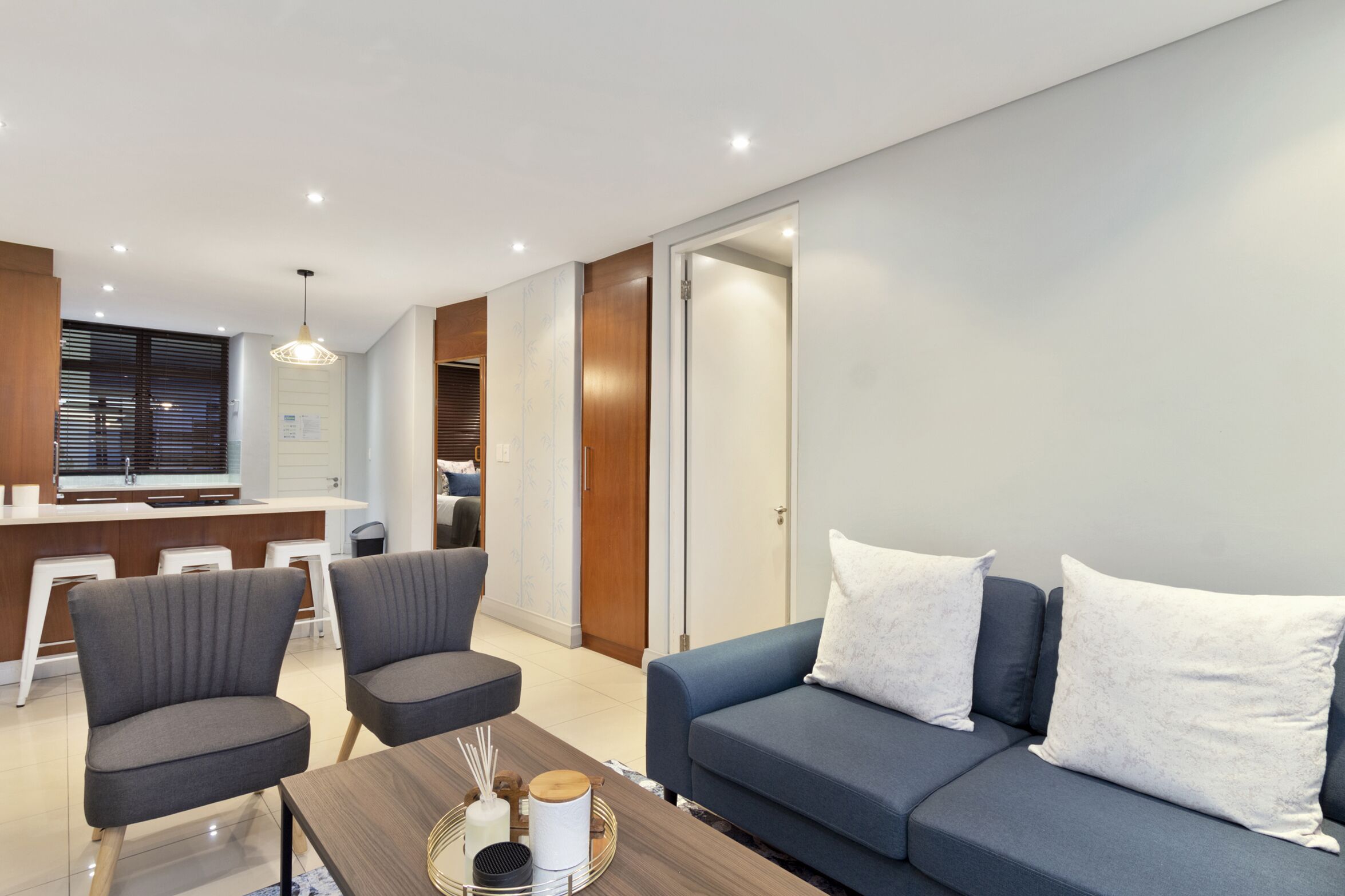 Uma sala de estar moderna com um sofá azul, duas poltronas cinzentas, uma mesa de centro de madeira e uma cozinha ao fundo.