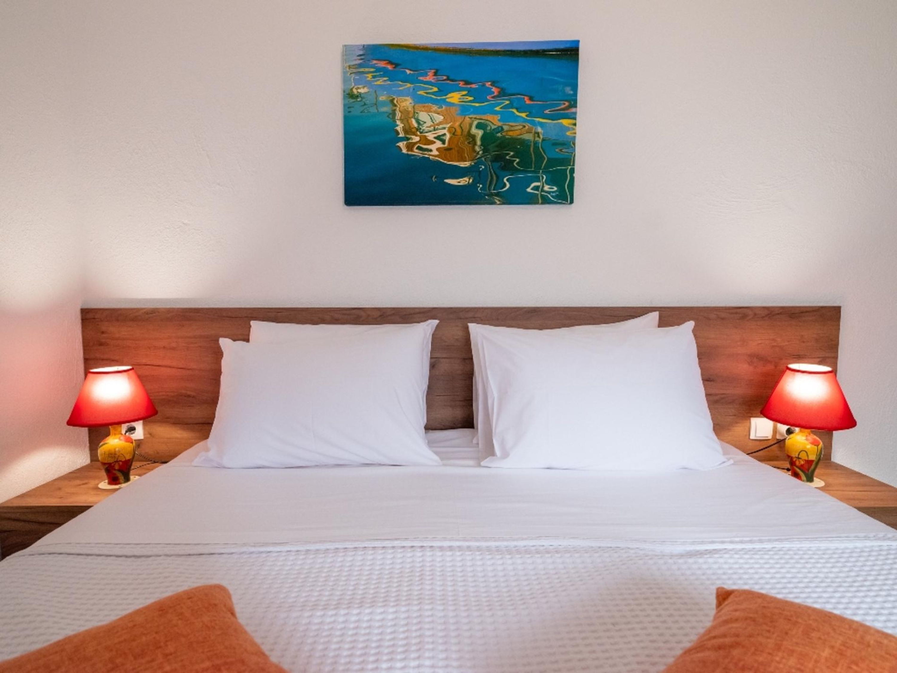 Cama bem arrumada com lençóis brancos e duas almofadas, com mesas de cabeceira de madeira com abajures vermelhos. Um quadro colorido na parede acima da cama.