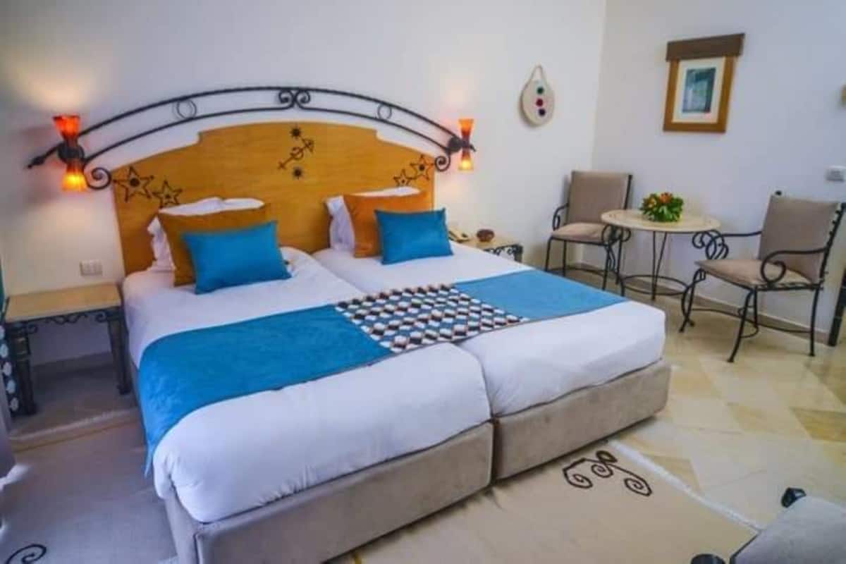 Uma cama de casal com cabeceira de madeira, dois travesseiros azuis e um cobertor xadrez. Há uma pequena mesa redonda com uma planta e duas cadeiras.