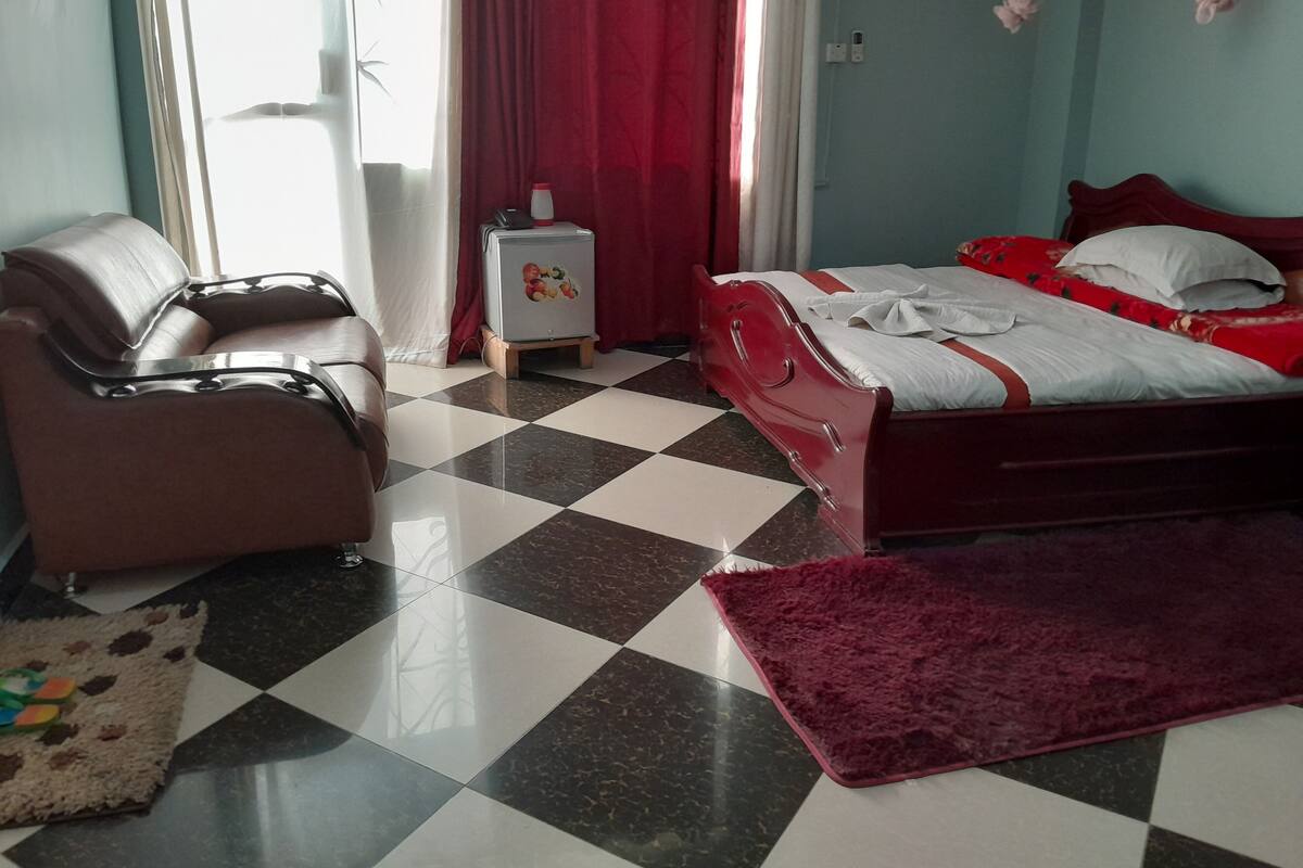 Quarto com piso xadrez, cama com roupa de cama vermelha, poltrona de couro marrom, armário branco pequeno e cortina vermelha.