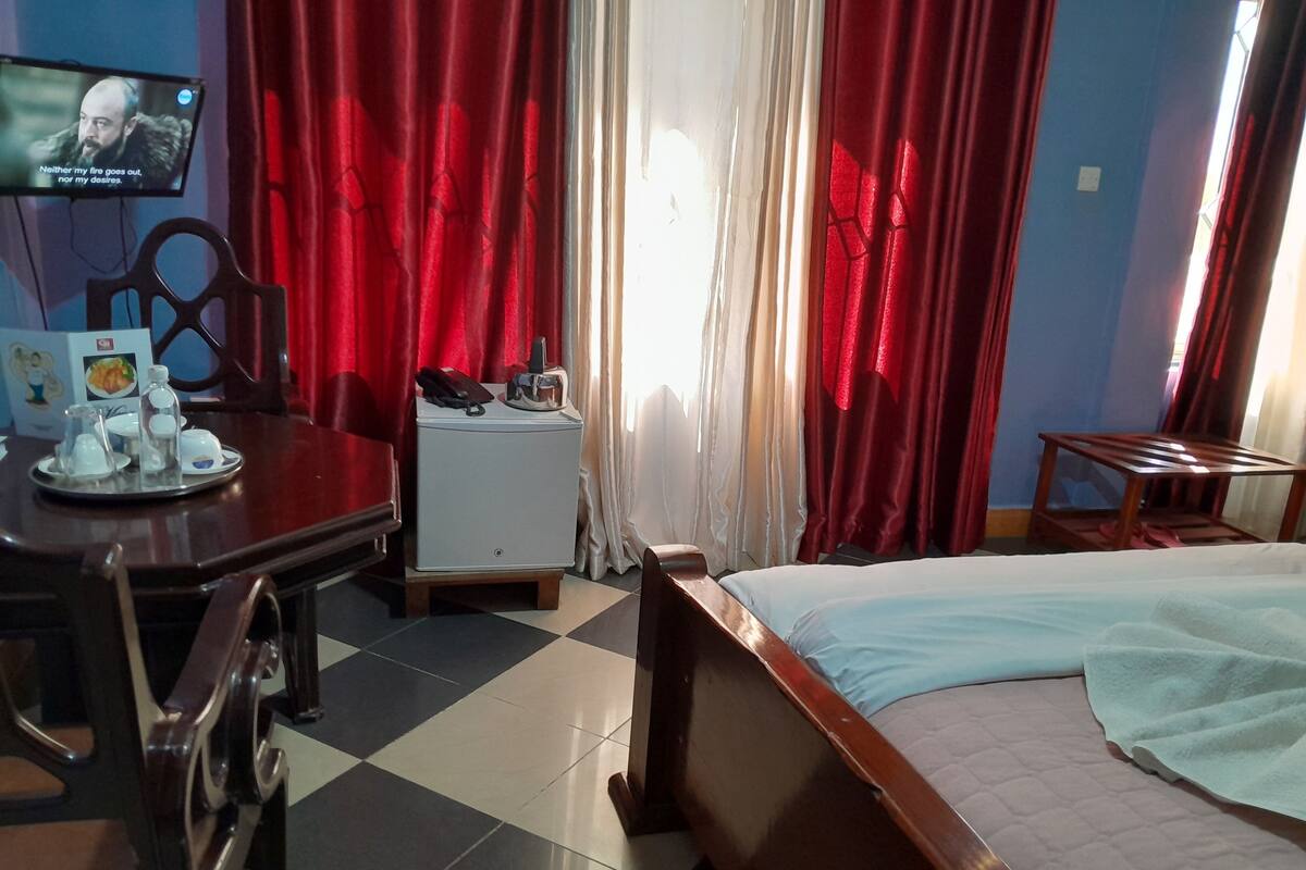Quarto de hotel com uma cama, uma escrivaninha com telefone e bandeja de chá, uma mesinha de apoio e televisão na parede.