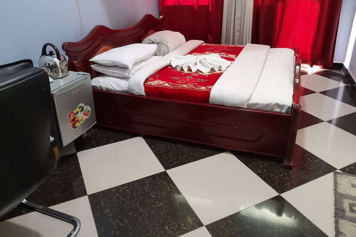 Quarto de hotel com piso xadrez, cama com colcha vermelha e branca, almofadas brancas e cabeceira de madeira. Há uma mesa pequena com uma chaleira e uma xícara, e uma cadeira com um desenho de frutas.