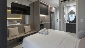 Quarto de hotel moderno com uma cama grande, um banco embutido, televisão de tela plana, minibar e banheiro com espelho e pia.