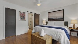 Quarto de hotel com cama, ventilador de teto, quadro na parede, mesa de cabeceira com abajur e porta para outro cômodo.
