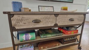 Um armário de madeira com duas gavetas e prateleiras, contendo jogos de tabuleiro, livros e um objeto decorativo azul.