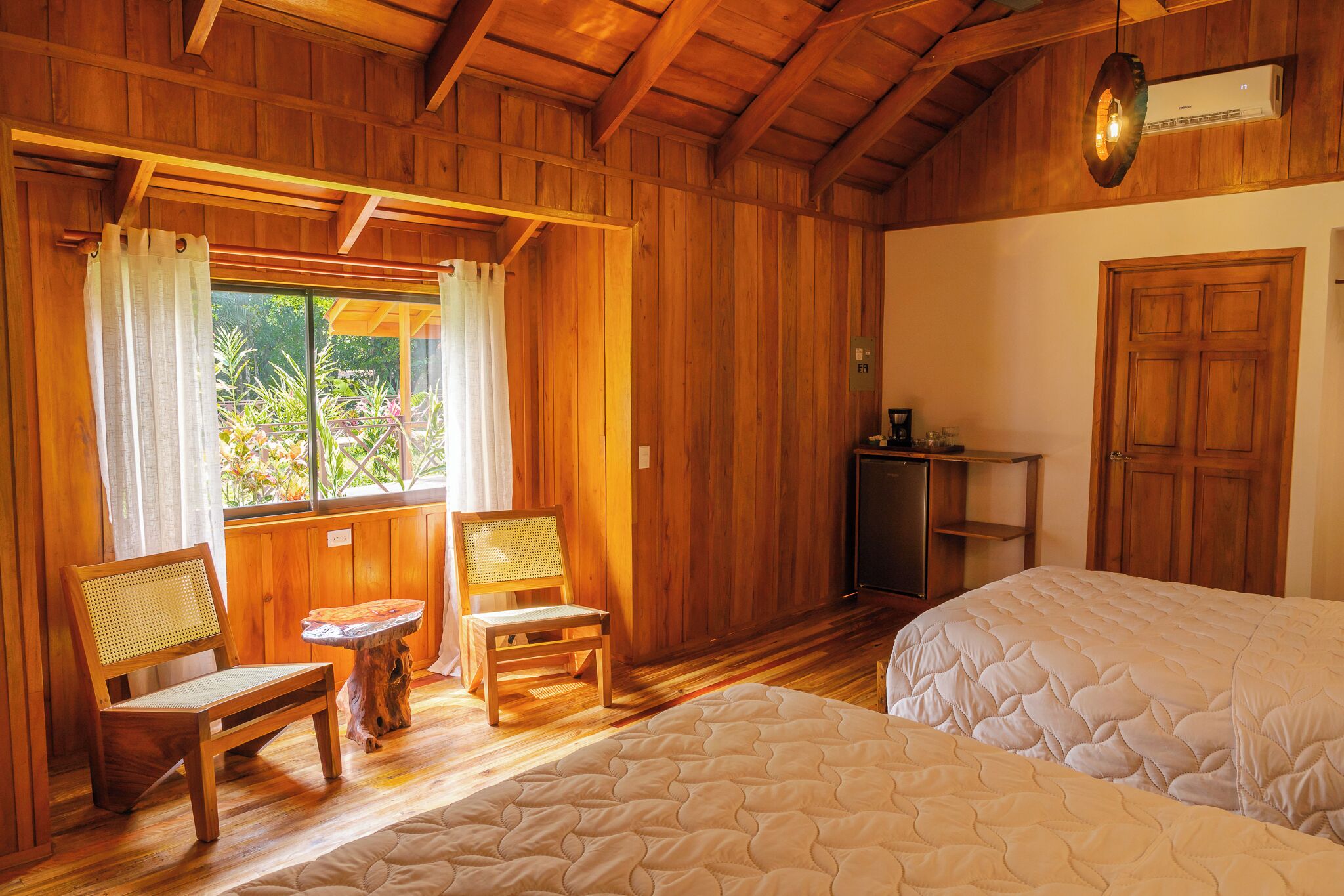 Um quarto de madeira com duas camas, uma cadeira de madeira, uma mesinha e uma janela com cortinas.