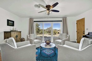 Quarto com ventilador de teto, duas poltronas, uma mesa de centro redonda, vista através de amplas janelas e uma porta de madeira.