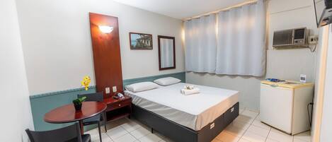 Quarto de hotel pequeno, com cama, mesa e cadeira, escrivaninha, geladeira e janela com cortinas.