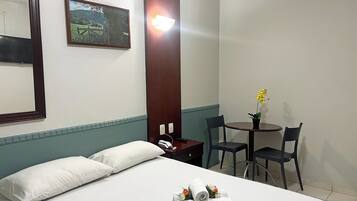 Quarto de hotel com cama, escrivaninha com telefone, cadeira, mesinha com planta e luminária na parede.