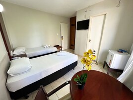 Quarto de hotel com duas camas, uma televisão, uma mesinha com uma planta e um frigorífico.