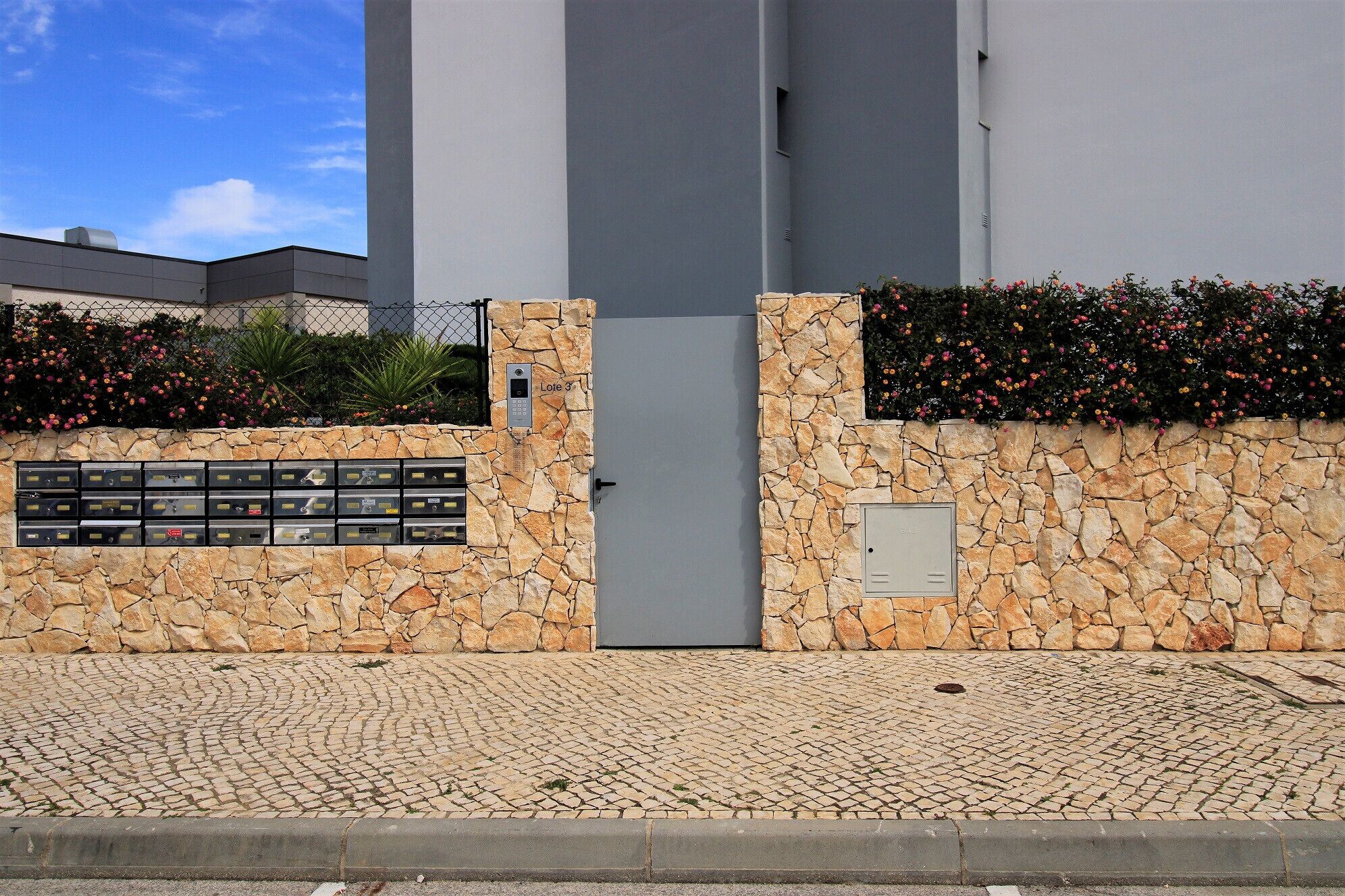 Entrada de um edifício com muro de pedra, portão de metal e calçada pavimentada.