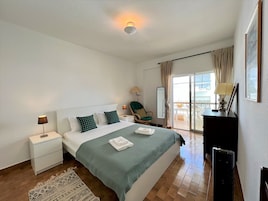 Quarto com cama, mesas de cabeceira, cadeira, escrivaninha e vista para o exterior.