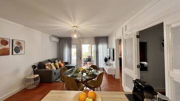 Uma sala de estar moderna com área de jantar, apresentando uma mesa de madeira com frutas, um sofá com almofadas e uma máquina de café.