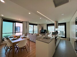 Uma cozinha moderna com uma ilha central, uma área de refeições com mesa e cadeiras, e uma sala de estar com sofá.