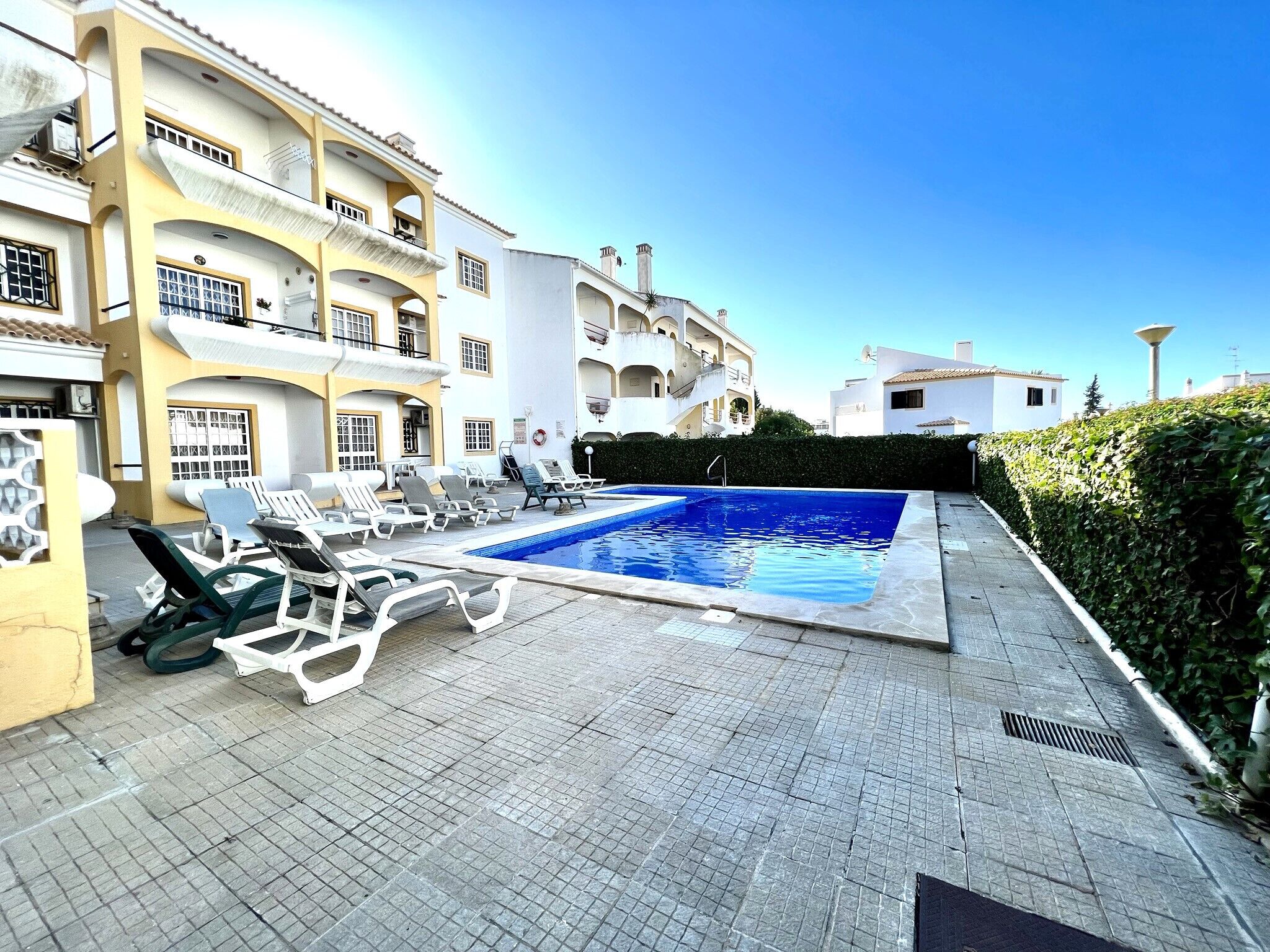 Uma área de piscina com espreguiçadeiras, um deck revestido de azulejos e um edifício com varandas.