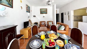 Uma área de refeições com uma mesa posta para o café da manhã, incluindo pratos, talheres e uma variedade de frutas e doces.