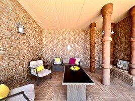 Quarto com parede de pedra, teto de madeira, mesa preta com um vaso, sofá cinza com almofadas coloridas e uma cadeira com almofada amarela.