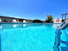 Uma piscina azul cristalina com um deck ladrilhado, cadeiras de lounge brancas e uma cerca de madeira ao fundo.