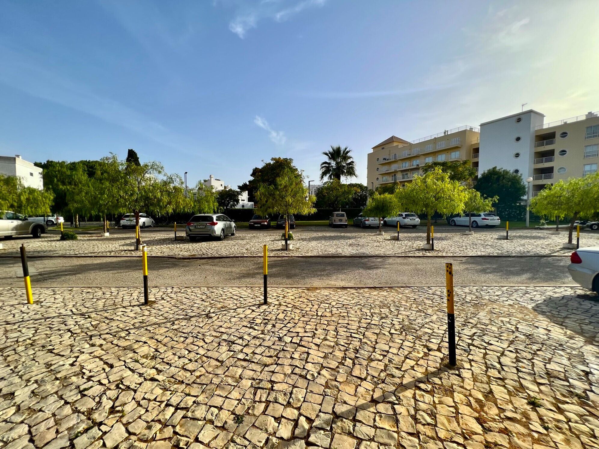 Uma área de estacionamento com calçada de pedras, vários carros estacionados e edifícios ao fundo.