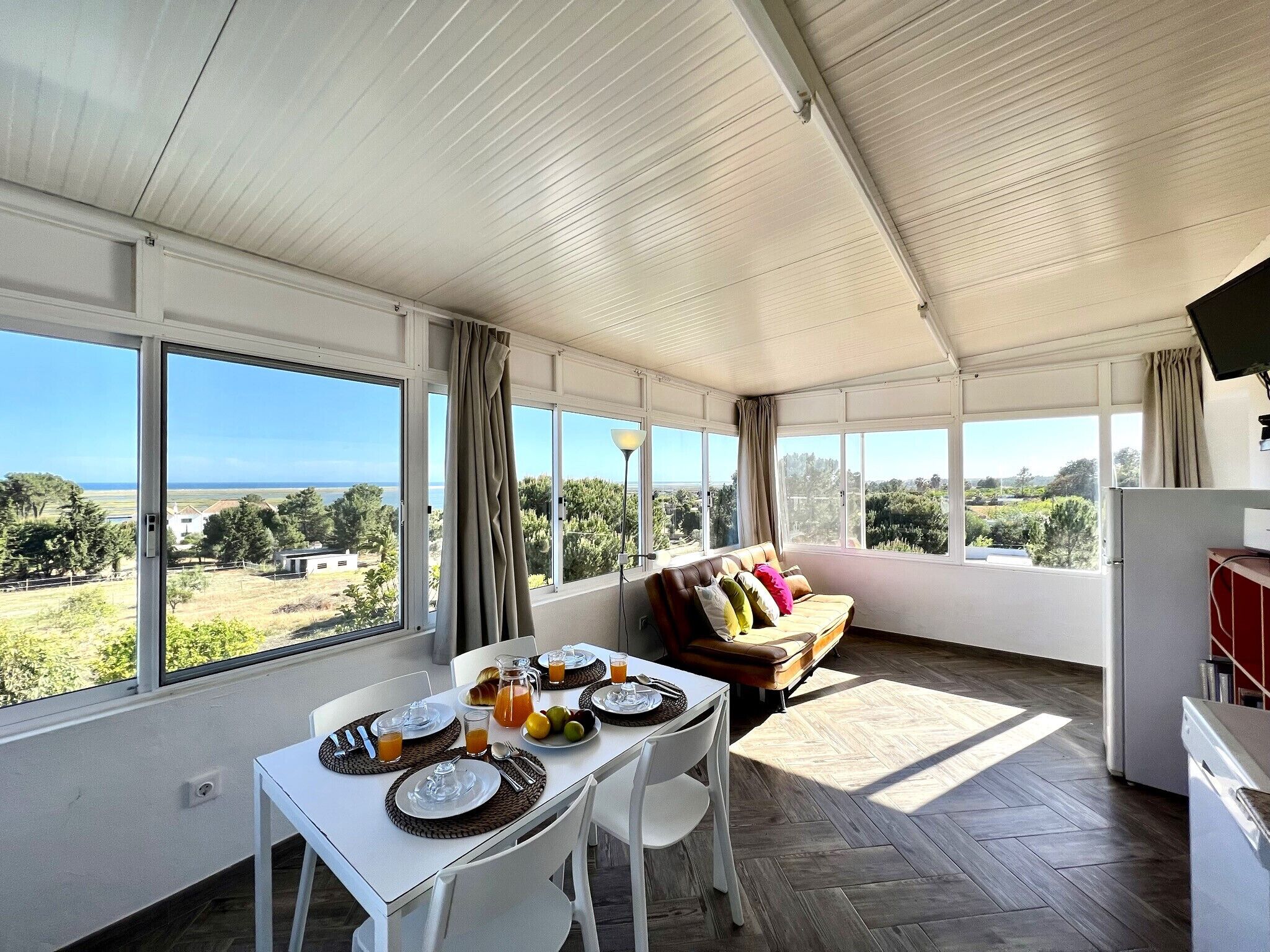 Um quarto iluminado pelo sol, com uma mesa de jantar posta para o café da manhã, um sofá com almofadas e grandes janelas com vista para o exterior.