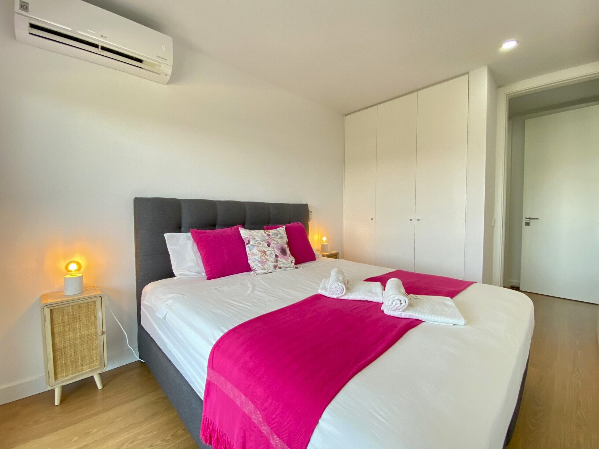 Uma cama bem arrumada com roupa de cama branca e rosa, uma cabeceira estofada cinza e uma mesa de cabeceira com um abajur.