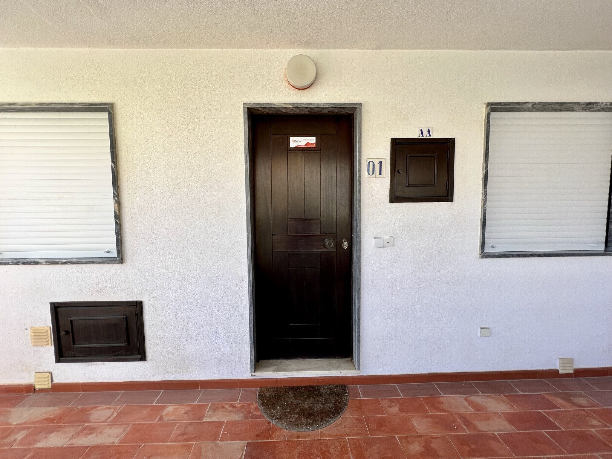 Entrada de um prédio com uma porta preta, duas janelas com persianas e uma placa com o número '01' acima da porta.