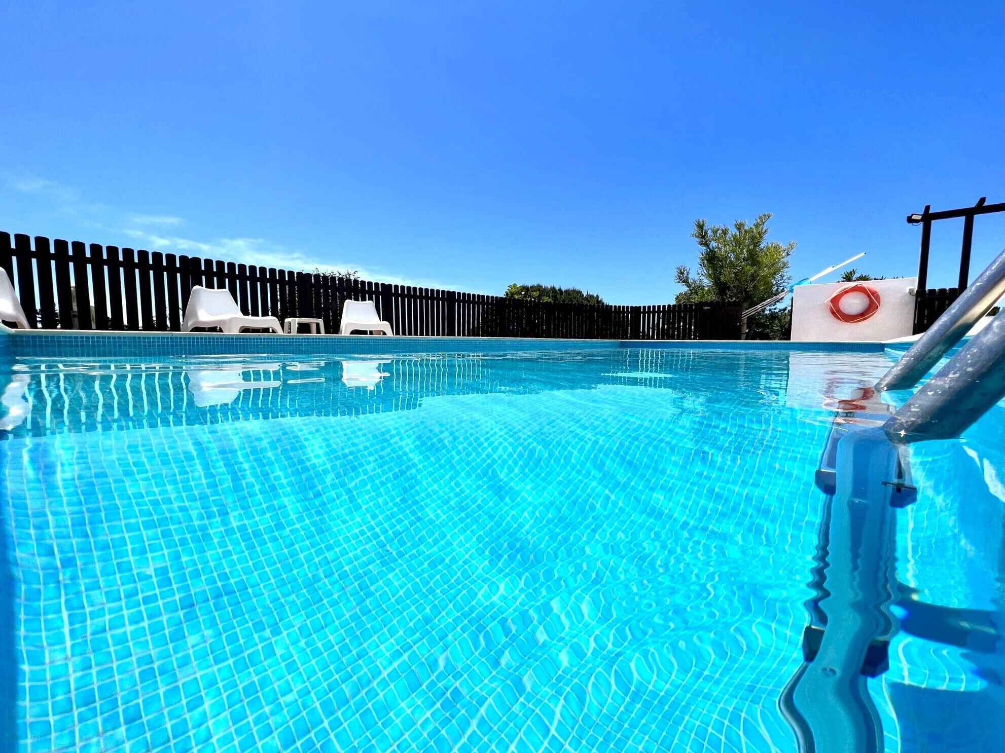 Uma piscina azul cristalina com um deck ladrilhado, cadeiras de lounge brancas e uma cerca de madeira ao fundo.