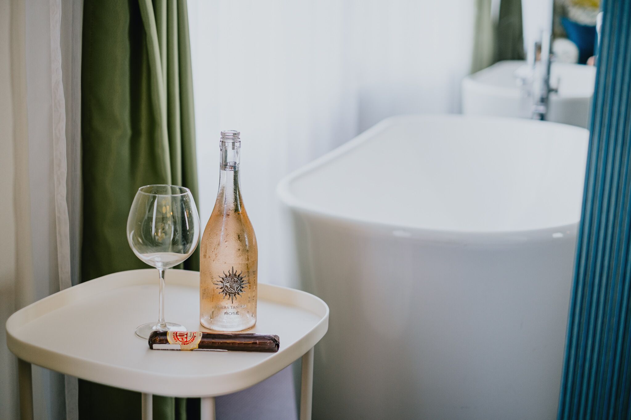 Uma garrafa de vinho rosé, um copo e um charuto sobre uma pequena mesa branca em um banheiro.