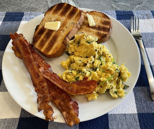 Um prato com duas fatias de pão torrado, ovos mexidos e uma fatia de bacon sobre uma toalha de mesa xadrez.