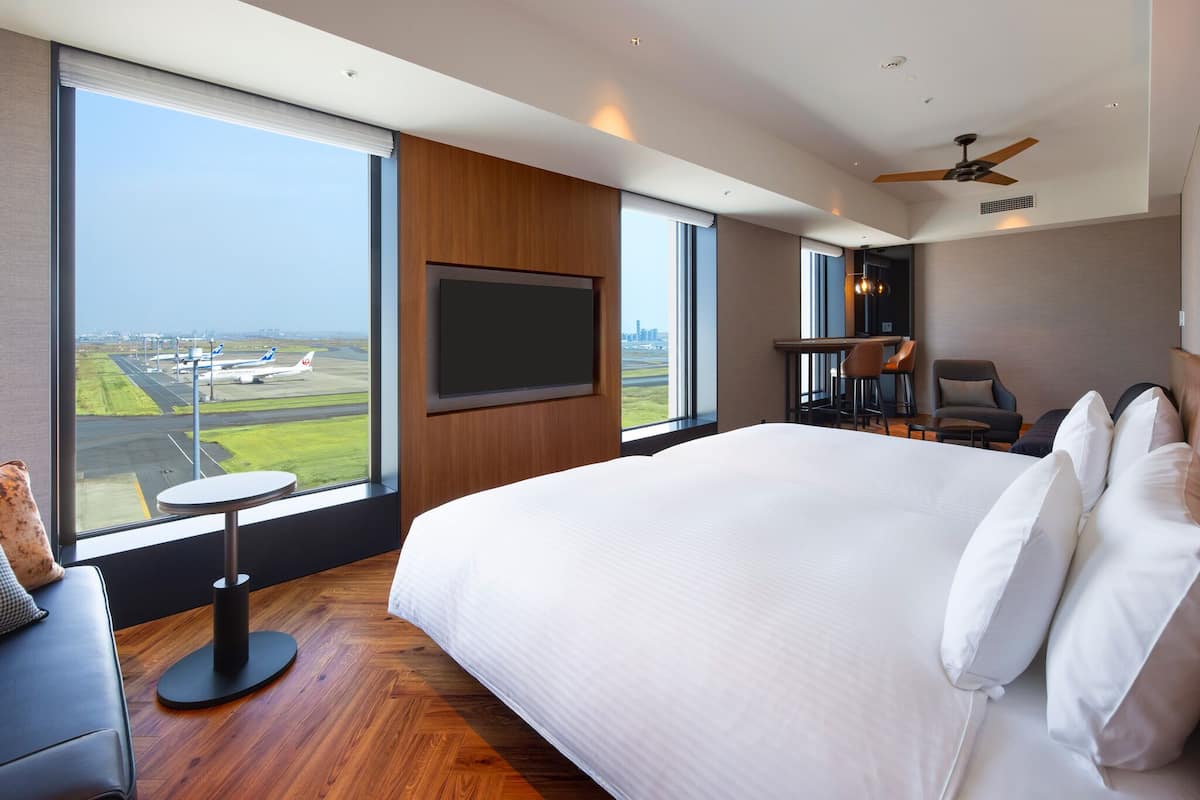 Quarto de hotel moderno com uma cama grande, uma mesa pequena, uma cadeira e uma TV fixada na parede.