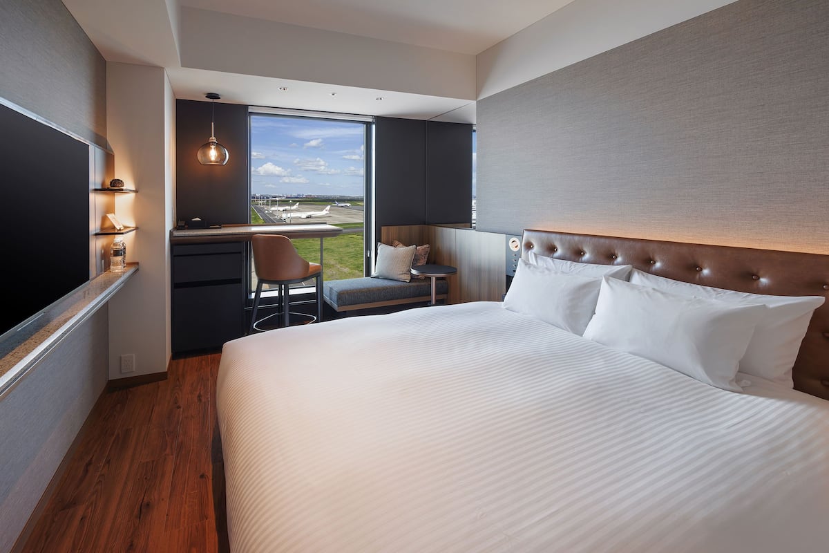 Quarto de hotel moderno com uma cama grande, uma escrivaninha e vista para o aeroporto.