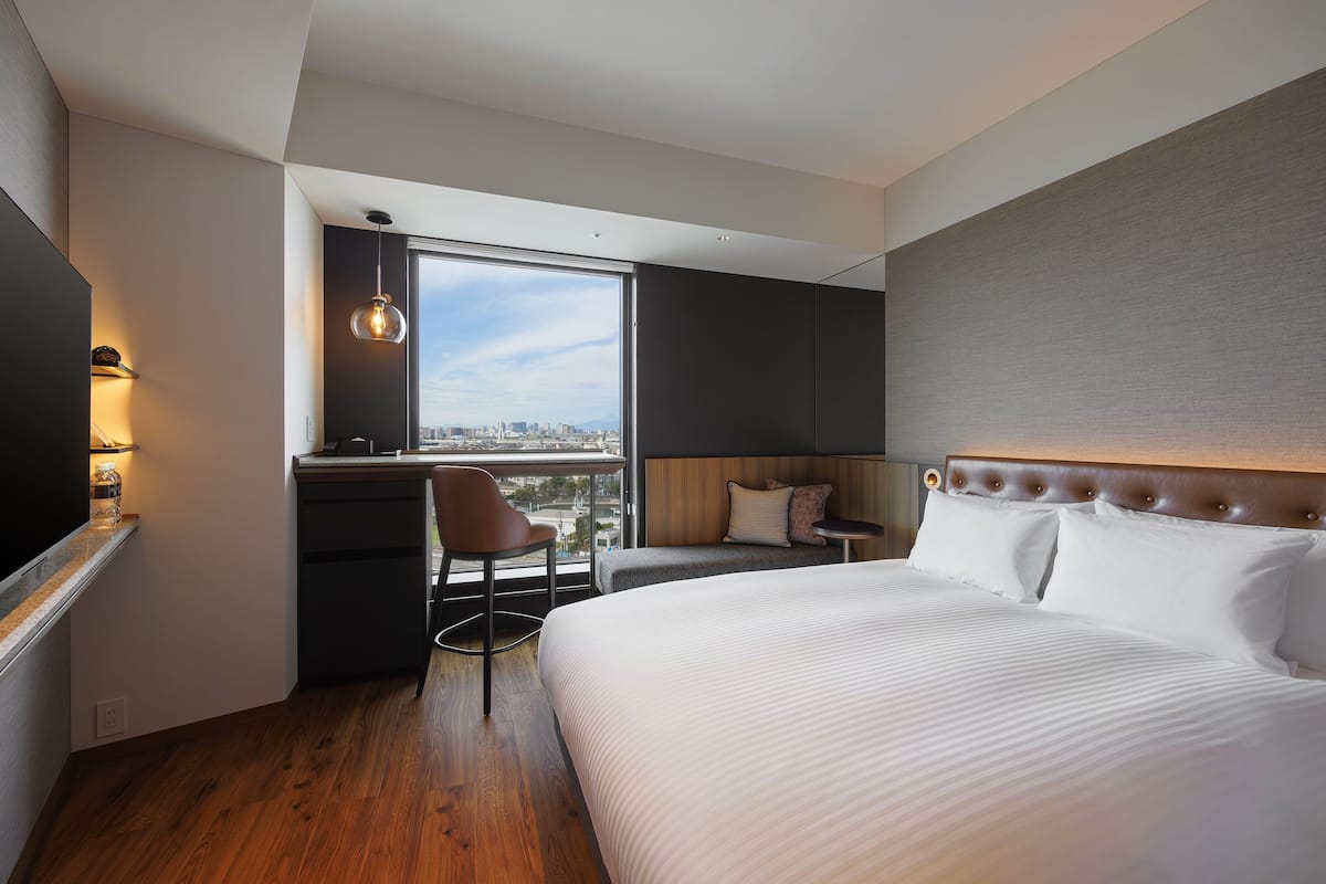 Quarto de hotel moderno com uma cama grande, uma escrivaninha com cadeira, uma janela com vista para a cidade e uma televisão à esquerda.