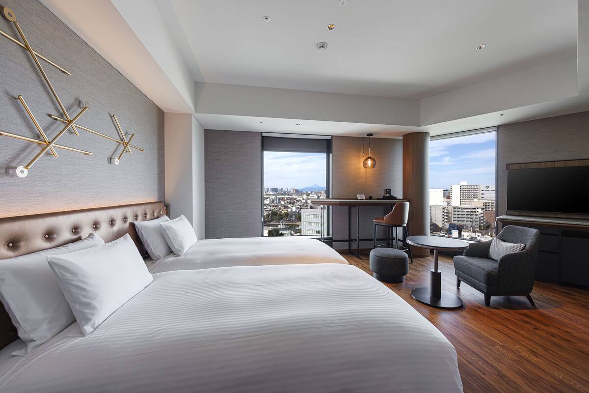 Quarto de hotel moderno com duas camas, um espaço de estar, televisão e vista para a cidade.