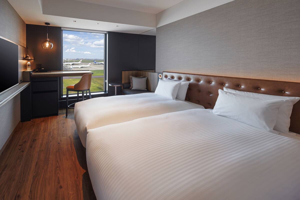Quarto de hotel moderno com uma cama grande, uma escrivaninha e vista para um aeroporto.