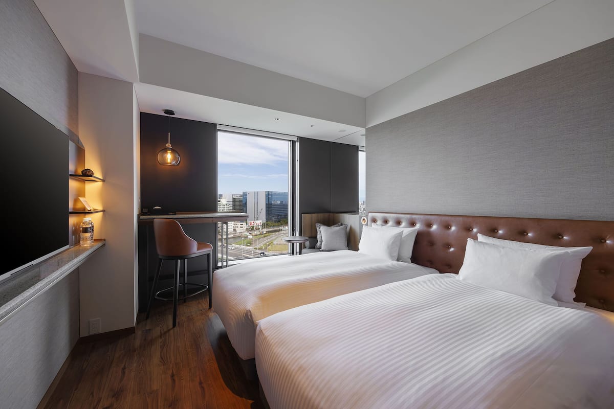 Quarto de hotel moderno com duas camas, uma janela ampla com vista para a cidade, uma pequena área de refeições e uma TV na parede esquerda.