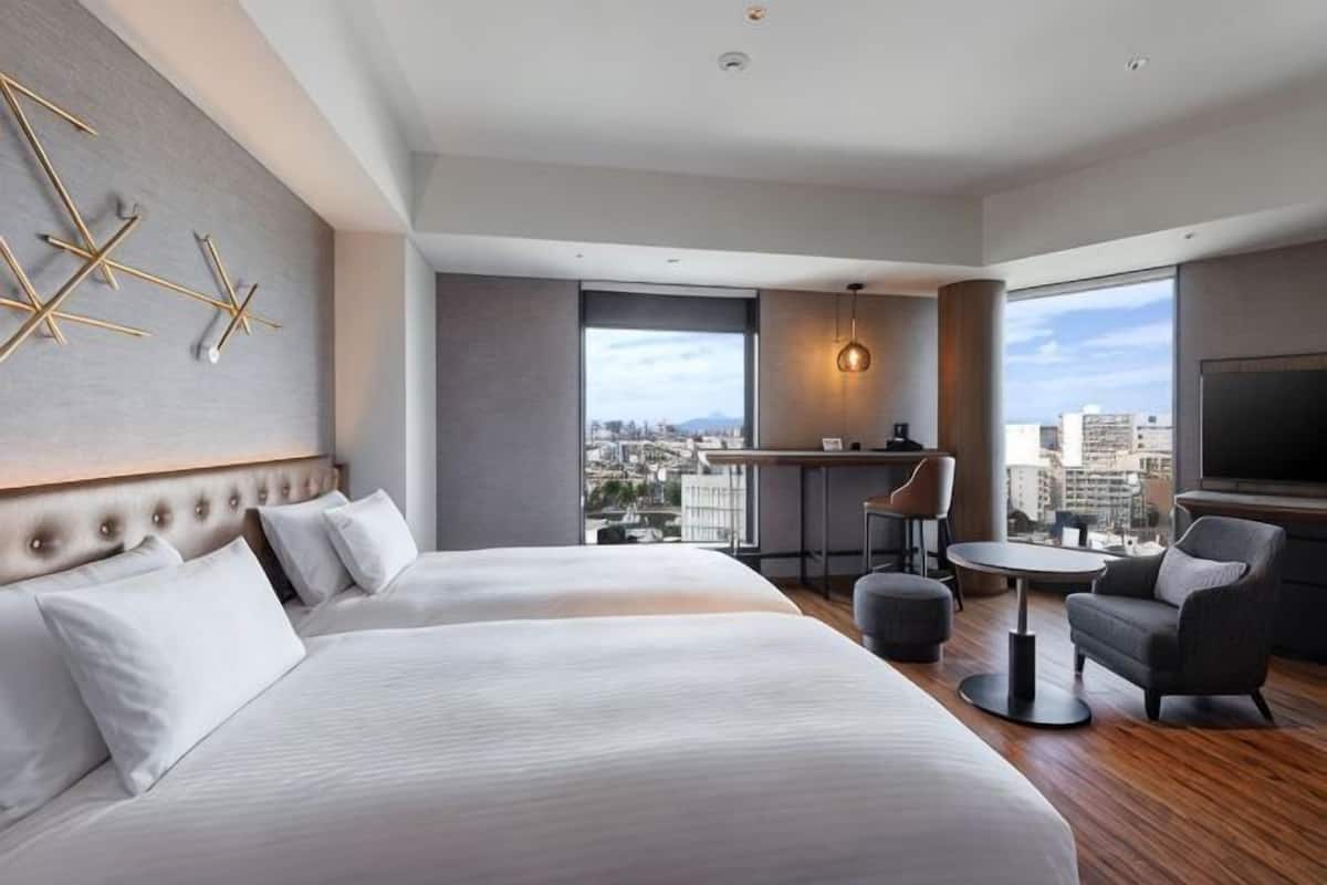 Quarto de hotel moderno com uma cama grande, uma pequena área de refeições e varanda com vista para a cidade.
