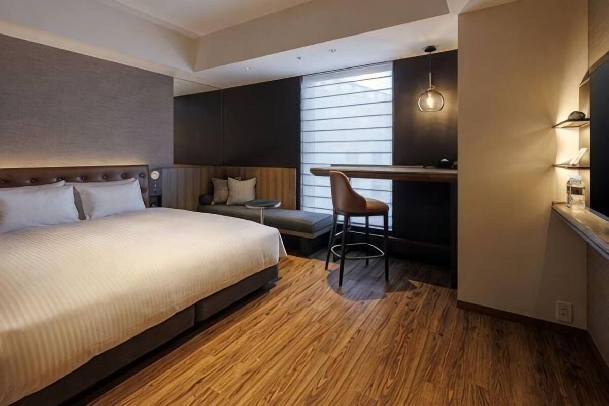 Quarto de hotel com uma cama grande, uma escrivaninha, uma cadeira e uma janela com persianas.