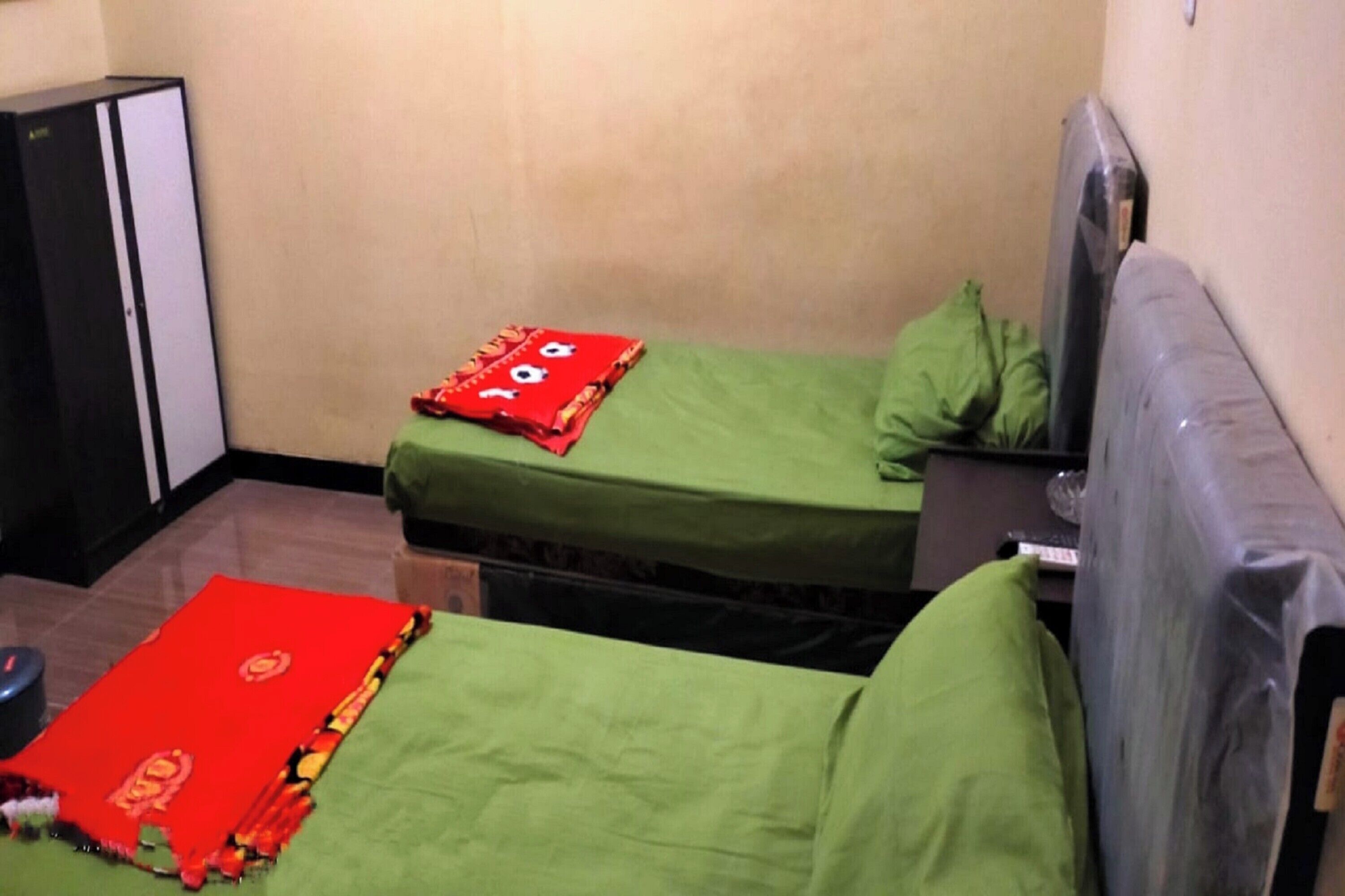 Quarto pequeno com duas camas, cada uma coberta por lençóis verdes e um cobertor vermelho. Há uma escrivaninha com uma cadeira e uma televisão sobre um suporte.