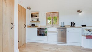 Uma cozinha moderna com armários brancos, forno embutido, lava-louças e uma janela com persiana florida.