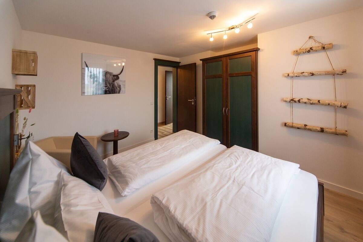 Quarto com duas camas, guarda-roupa de madeira, mesa pequena e prateleira na parede.