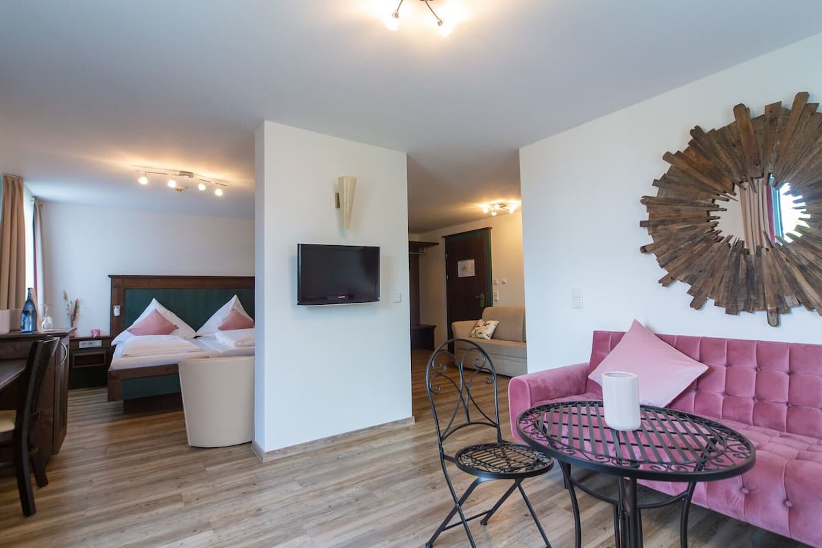 Quarto de hotel com cama, televisão, sofá rosa, mesa redonda e um objeto decorativo na parede.