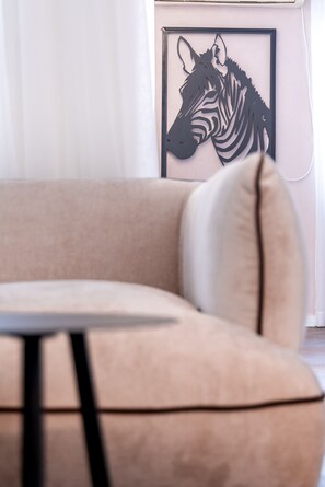 Um sofá bege com uma mesa redonda na frente, uma obra de arte com zebras emoldurada na parede e uma cortina branca.