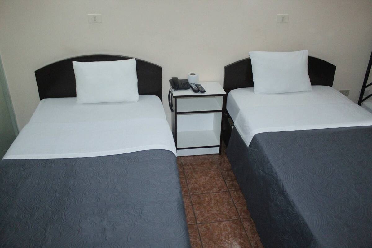 Quarto de hotel com duas camas de solteiro, cada uma com almofada branca e cobre escura. Há uma pequena mesa de cabeceira com um telefone, controles remotos e um rolo de papel higiênico.